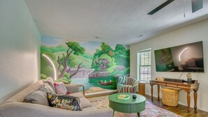 Uma sala de estar com um mural, um sofá, uma mesa de centro, uma televisão e uma janela.