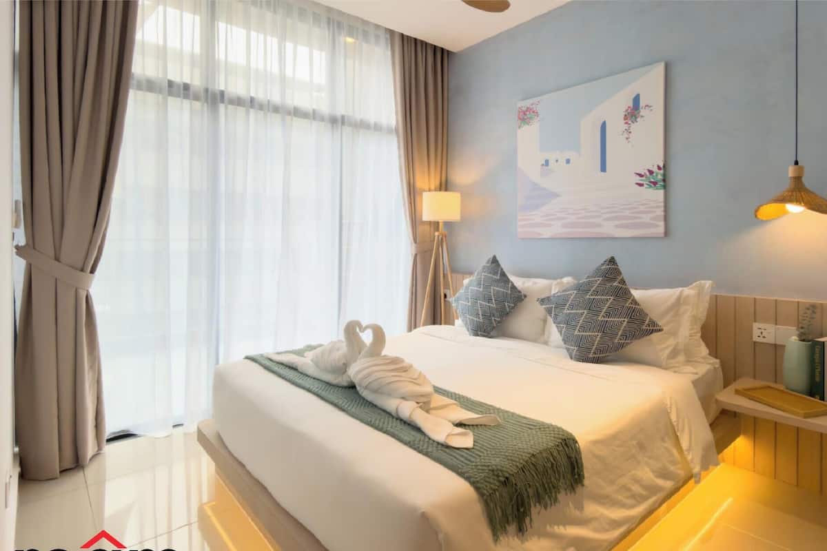 Quarto de hotel com cama, cortinas, luminária, pintura e mesa de cabeceira.