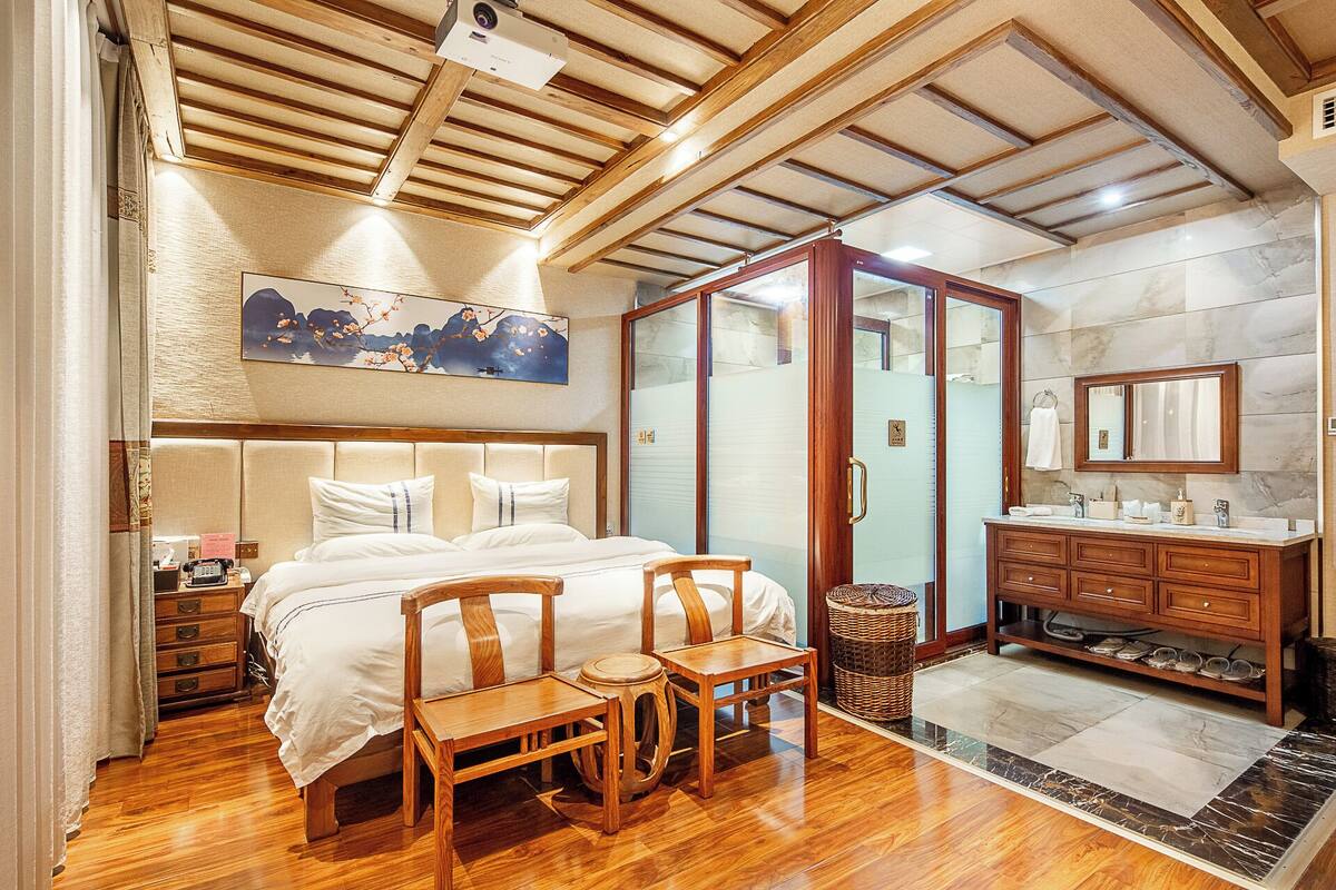 Quarto de hotel com teto de madeira, uma cama grande com roupa de cama branca, uma cadeira de madeira, uma penteadeira de madeira com espelho e uma janela ampla com vista para árvores.