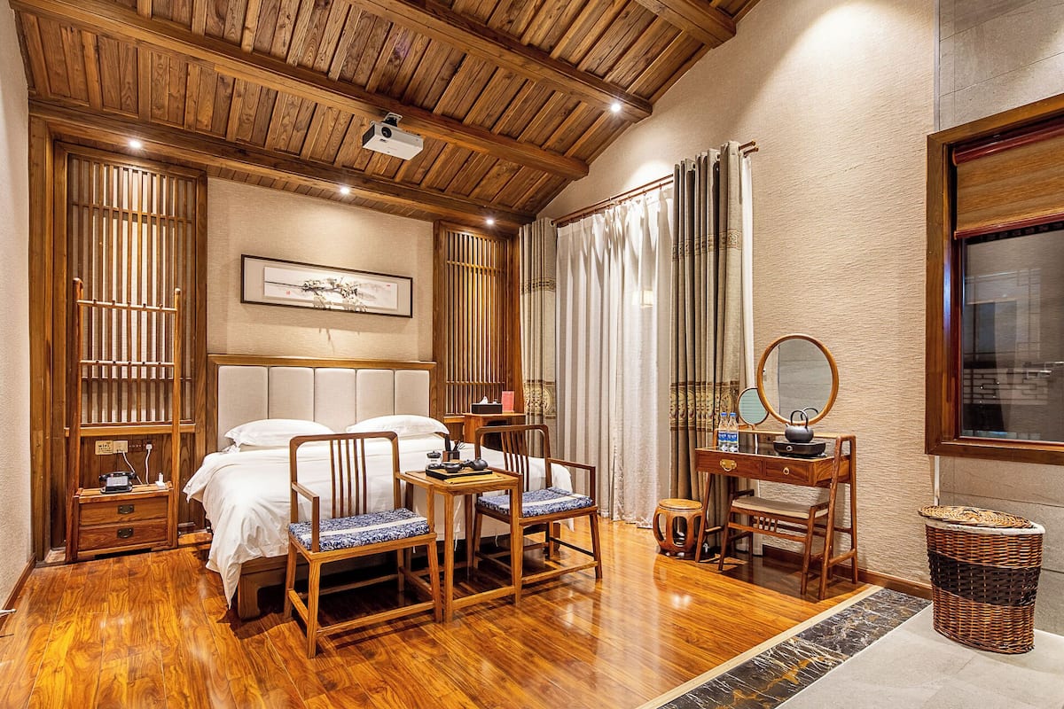 Quarto de hotel com teto de madeira, cama com lençóis brancos, duas cadeiras de madeira, uma mesa pequena, um espelho, um cesto de vime e uma janela com cortinas.