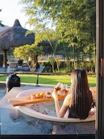 Mulher sentada à mesa junto à piscina, com uma bandeja de comida e bebidas, em meio a um jardim exuberante e uma estrutura com telhado de palha.
