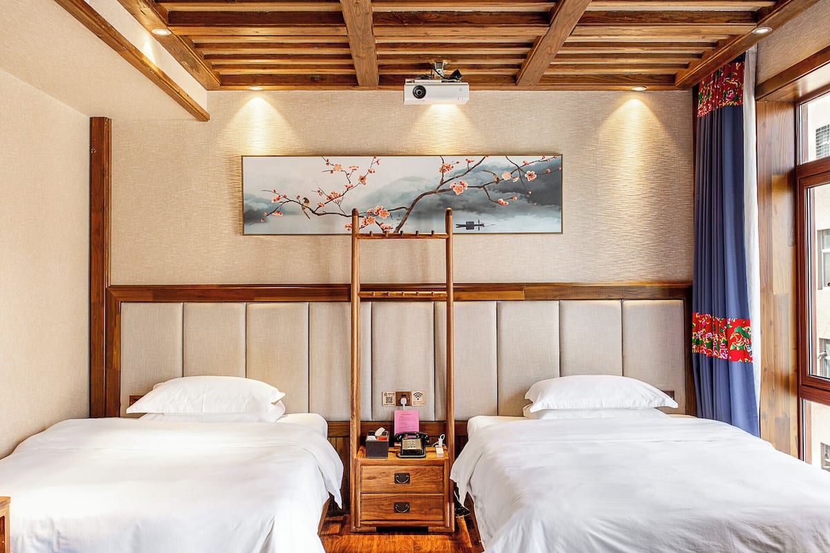 Quarto de hotel com duas camas, teto de madeira, pintura de um galho e janela com cortinas.