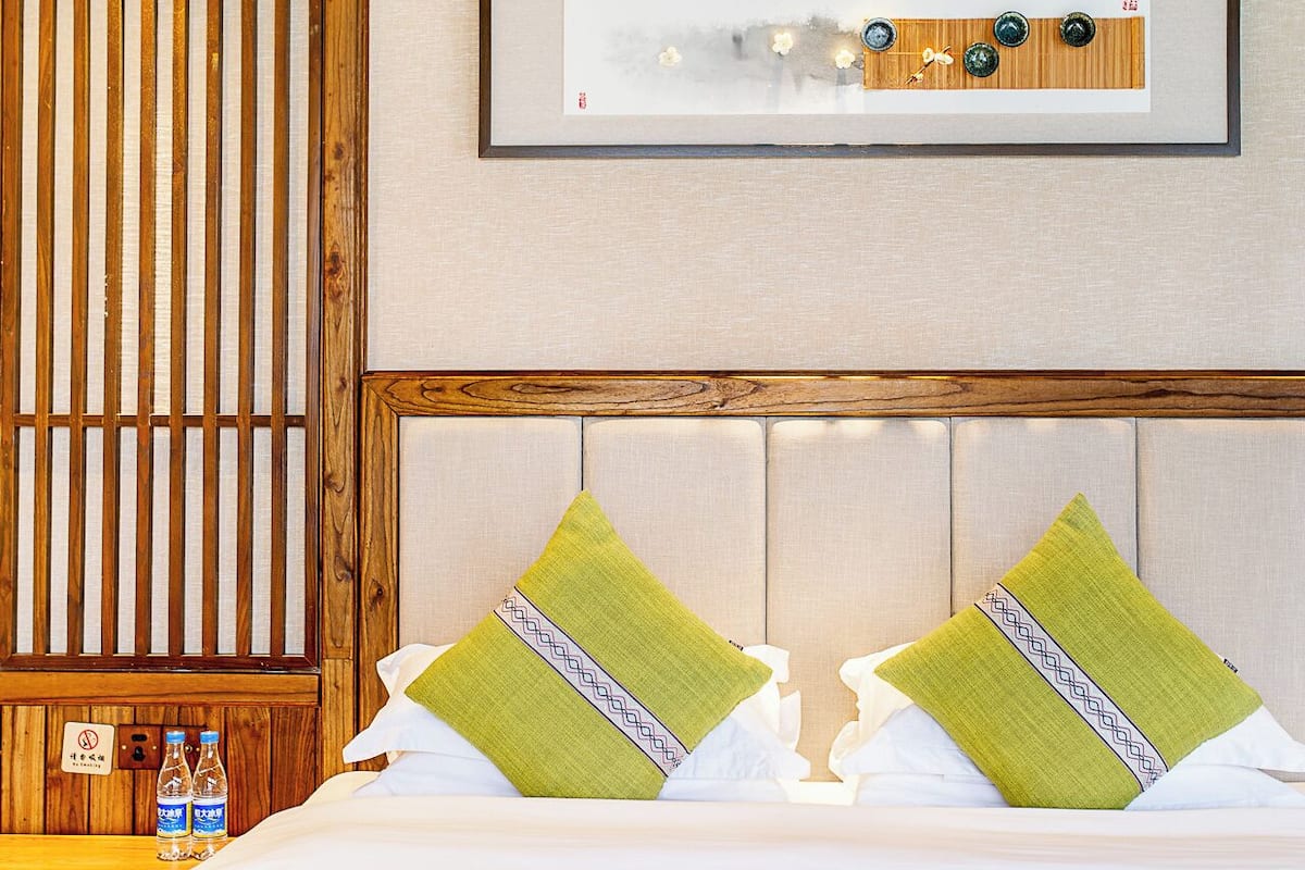 Quarto de hotel com teto de madeira, cama com almofadas verdes, espelho fixado na parede e mesa de cabeceira com garrafas de água.
