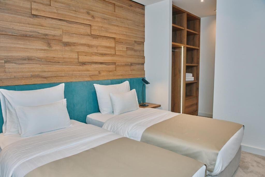 Quarto de hotel com duas camas, cabeceira de madeira, mesa de cabeceira e closet espaçoso.