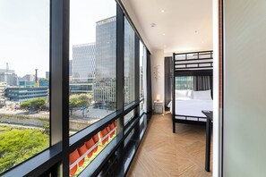 Quarto de hotel moderno com uma janela grande, uma cama, uma mesinha de cabeceira e vista para a paisagem urbana.