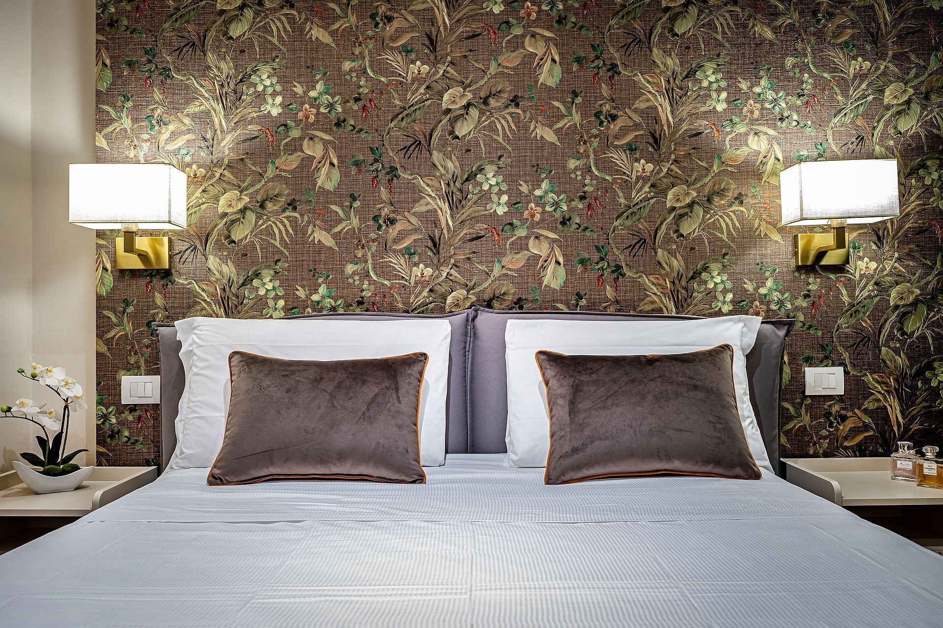 Quarto de hotel com papel de parede floral, duas luminárias de cabeceira, cama com lençóis brancos e mesa de cabeceira com uma garrafa.