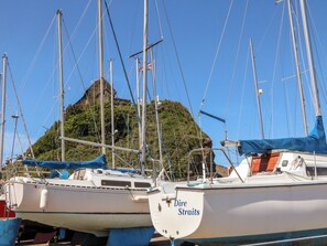 Uma marina com vários veleiros atracados, um edifício de pedra ao fundo e uma colina coberta de vegetação.