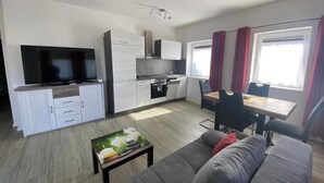 Uma sala de estar moderna com sofá, televisão, mesa de jantar com cadeiras, área de cozinha e janela com cortinas.