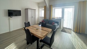 Uma sala de estar moderna com uma mesa de jantar de madeira, cadeiras pretas, um sofá cinza com almofadas, uma TV de tela plana na parede e um guarda-roupa.
