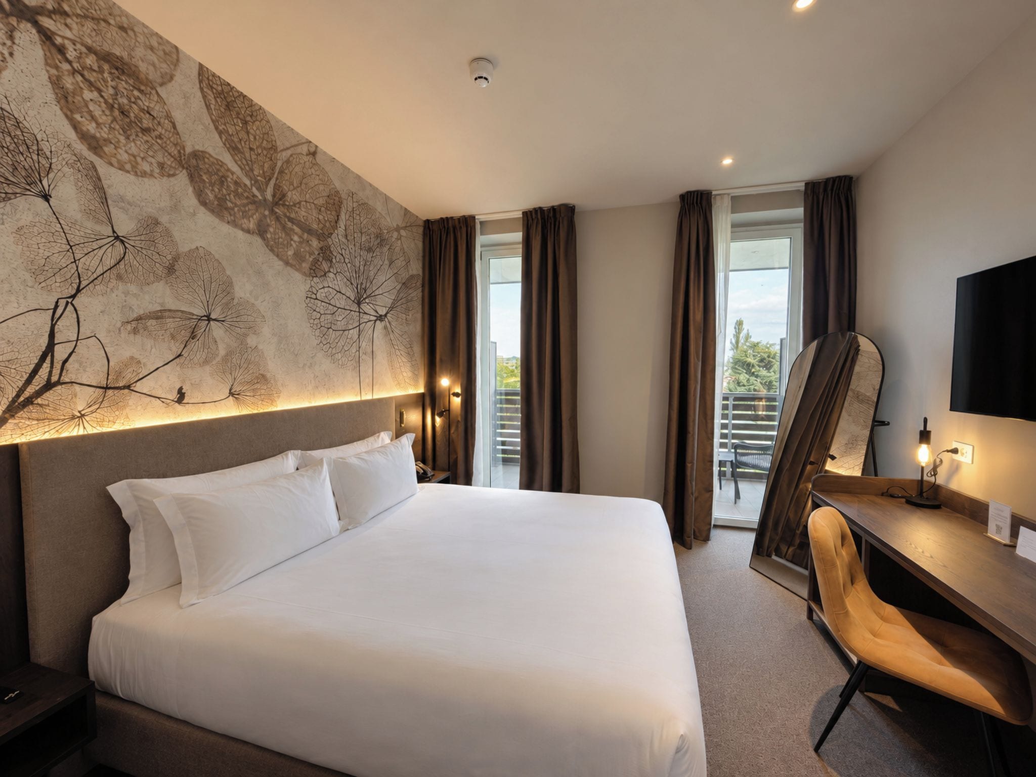 Quarto de hotel com uma cama grande, uma escrivaninha com cadeira, televisão de tela plana e vista para o exterior pelas janelas.