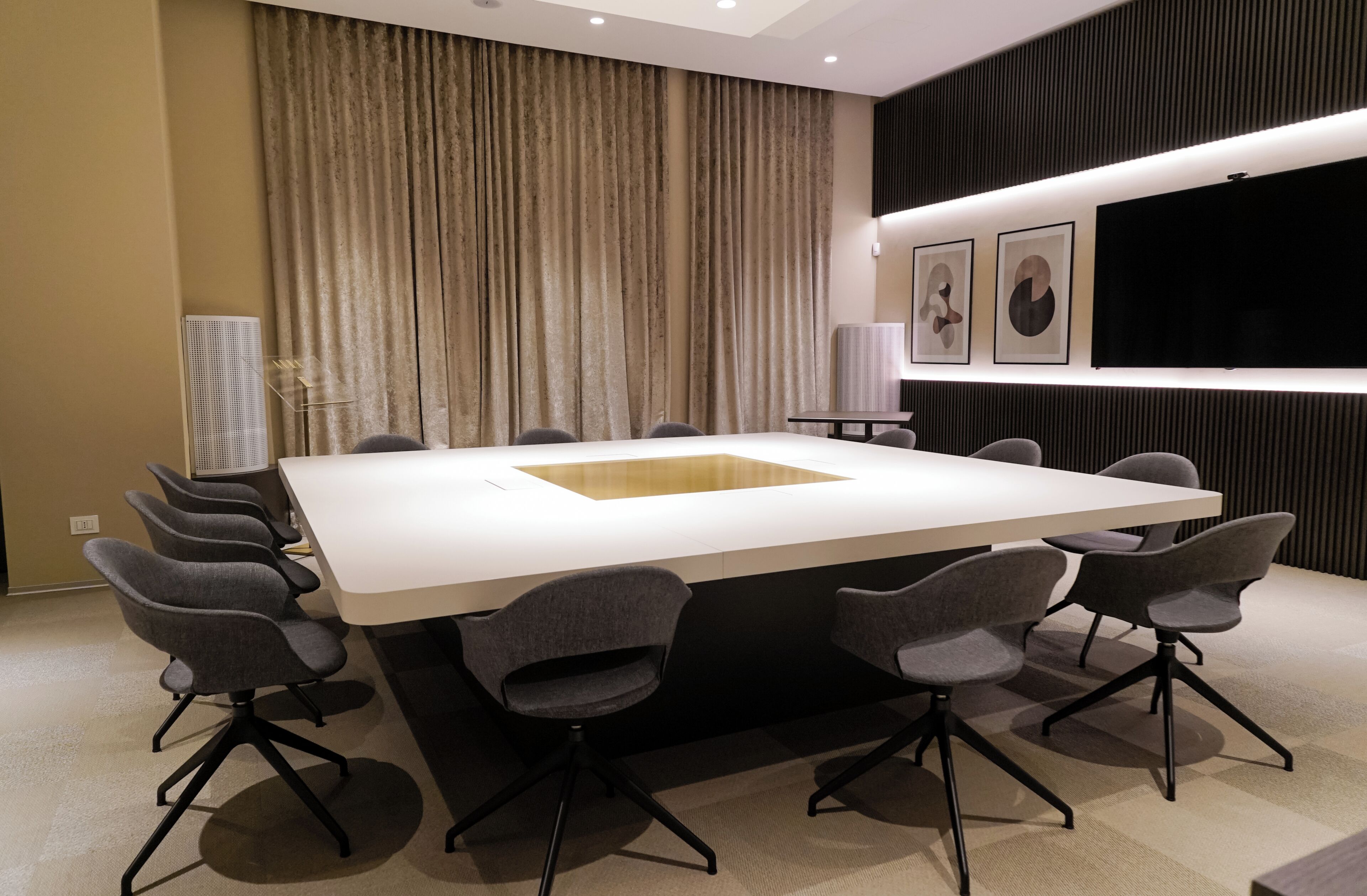 Uma sala de conferências com uma mesa grande, cadeiras cinzentas, uma cortina, um abajur e quadros na parede.