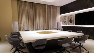 Uma sala de conferências com uma mesa grande, cadeiras cinzentas, uma cortina, um abajur e quadros na parede.