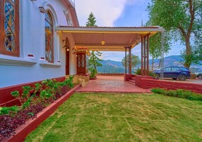Um edifício com entrada coberta, um jardim com plantas e vista para as montanhas.
