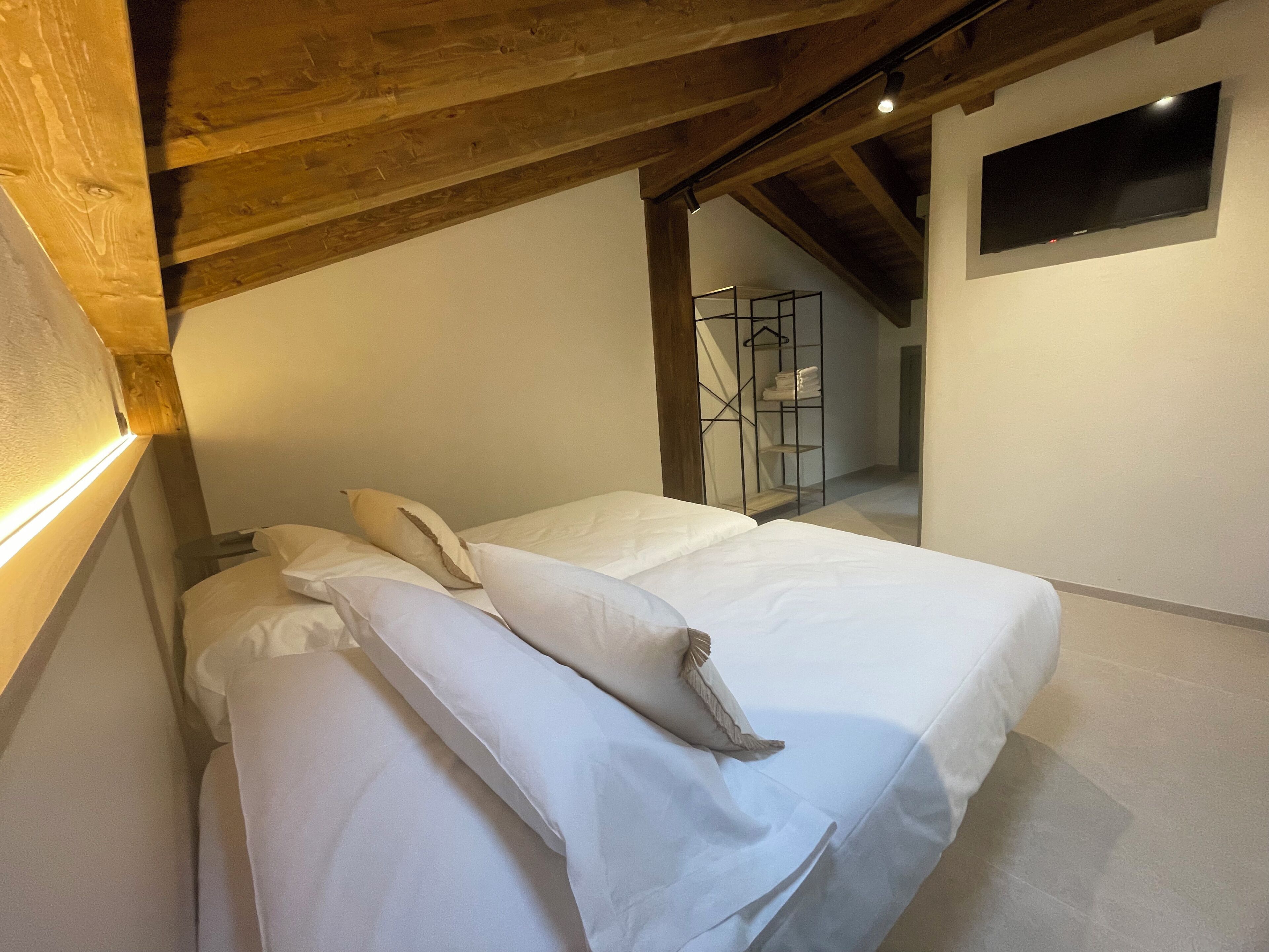 Quarto com teto de madeira, cama com roupa de cama branca, televisão fixada na parede e corredor com estante de metal.