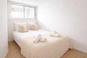 Uma cama de solteiro com um cobre-leito bege, almofadas brancas e toalhas enroladas sobre um piso de madeira.