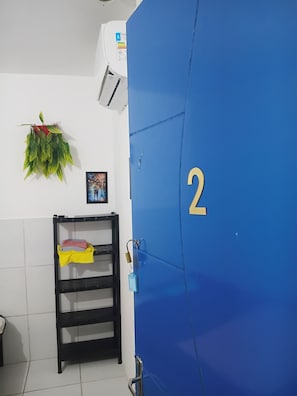 Quarto com porta azul numerada 2, prateleira pequena com roupas dobradas, ar-condicionado fixado na parede e planta em vaso.