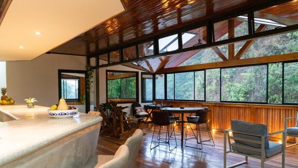 Um quarto espaçoso com teto de madeira, janelas amplas, área de jantar com cadeiras e mesa, e bancada de cozinha com frutas.