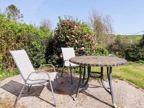 Mesa redonda com duas cadeiras, cercada por um jardim com um arbusto de flores cor-de-rosa.
