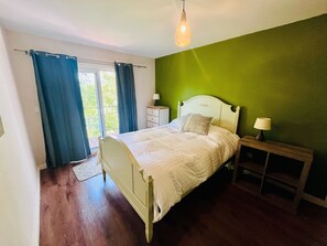 Quarto com cama, mesa de cabeceira, luminária, janela com cortinas e vista para o verde lá fora.