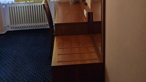 Quarto de hotel com uma secretária de madeira, uma cadeira, uma televisão, um abajur, um radiador e uma janela com cortinas amarelas.