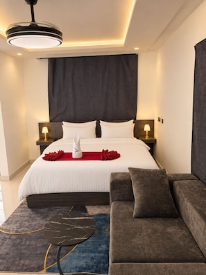 Quarto de hotel moderno com uma cama grande, duas mesas de cabeceira com luminárias, um painel de cabeceira escuro, uma toalha vermelha, um sofá cinza e um tapete azul estampado.