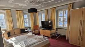 Quarto de hotel com cama, televisão, sofá, poltrona, mesa pequena e guarda-roupa.