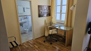 Quarto com piso de madeira, uma escrivaninha com cadeira, janela com cortinas, espelho e uma cadeira de rodas.
