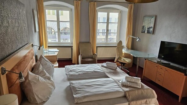 Quarto de hotel com cama, escrivaninha, cadeira, televisão e janelas amplas com cortinas.