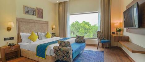 Quarto de hotel com uma cama grande, mesas de cabeceira, uma escrivaninha, uma cadeira, uma televisão e uma janela com vista para árvores.