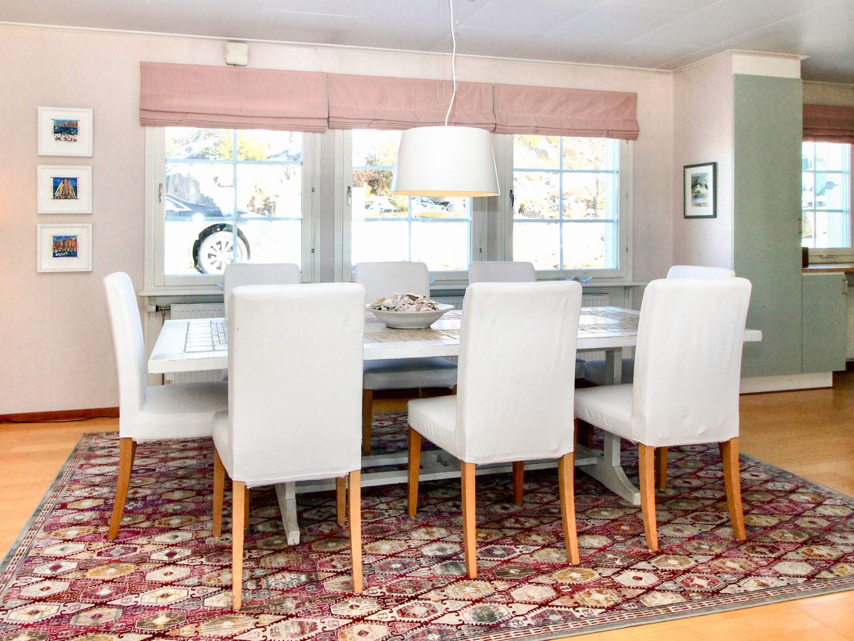 Uma sala de jantar com uma mesa branca, quatro cadeiras, um tapete estampado e uma janela com vista para um carro e árvores.