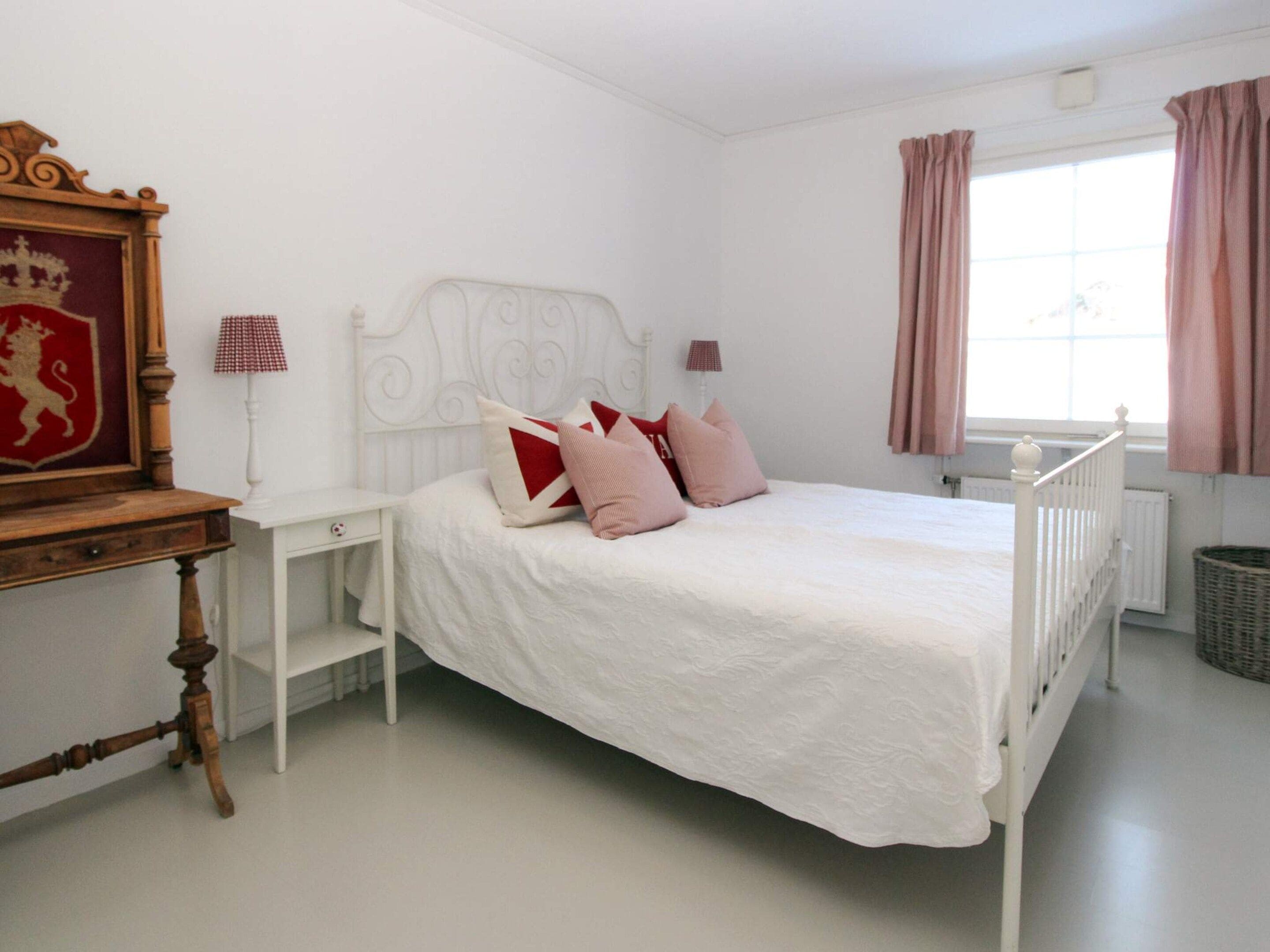 Quarto com cama branca, escrivaninha de madeira, quadro com moldura vermelha e branca e janela com cortinas.