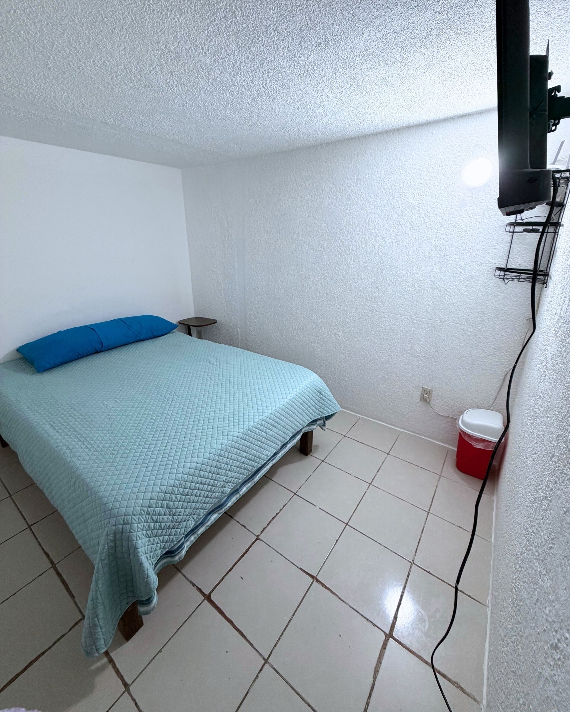 Um quarto pequeno e pouco mobiliado, com uma cama, uma mesinha e um balde vermelho.