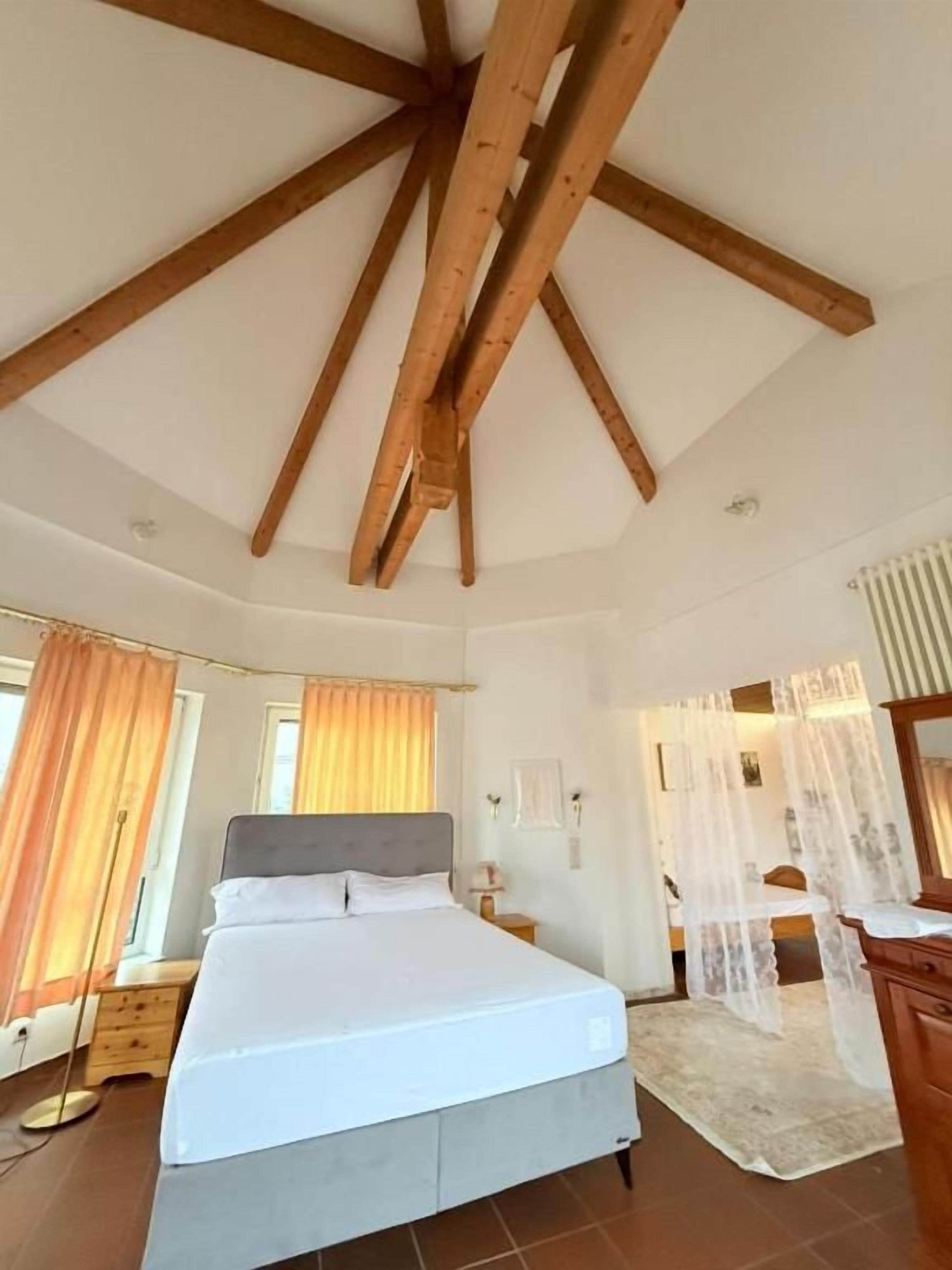 Quarto com cama, vigas de madeira, mesa de cabeceira, cômoda e janela com cortinas.