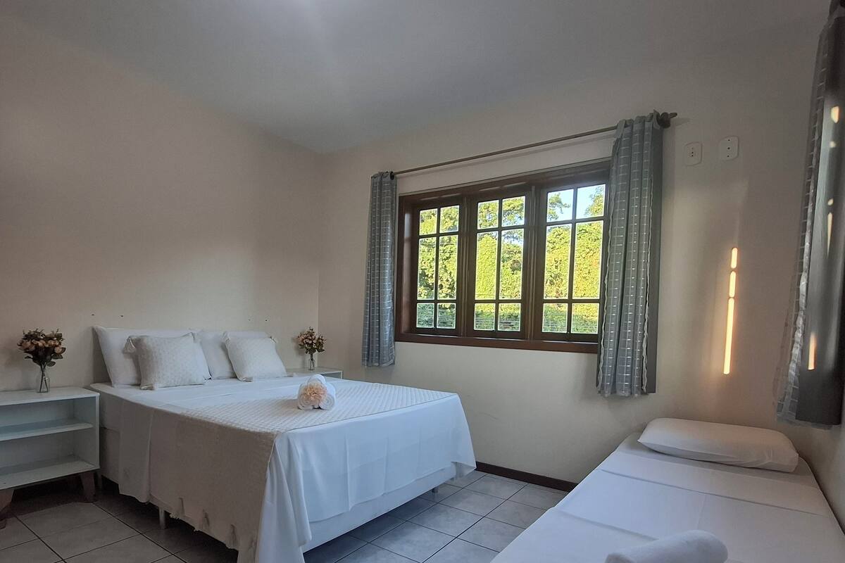 Quarto com duas camas, janela com cortinas, mesa de cabeceira com um vaso e piso ladrilhado.