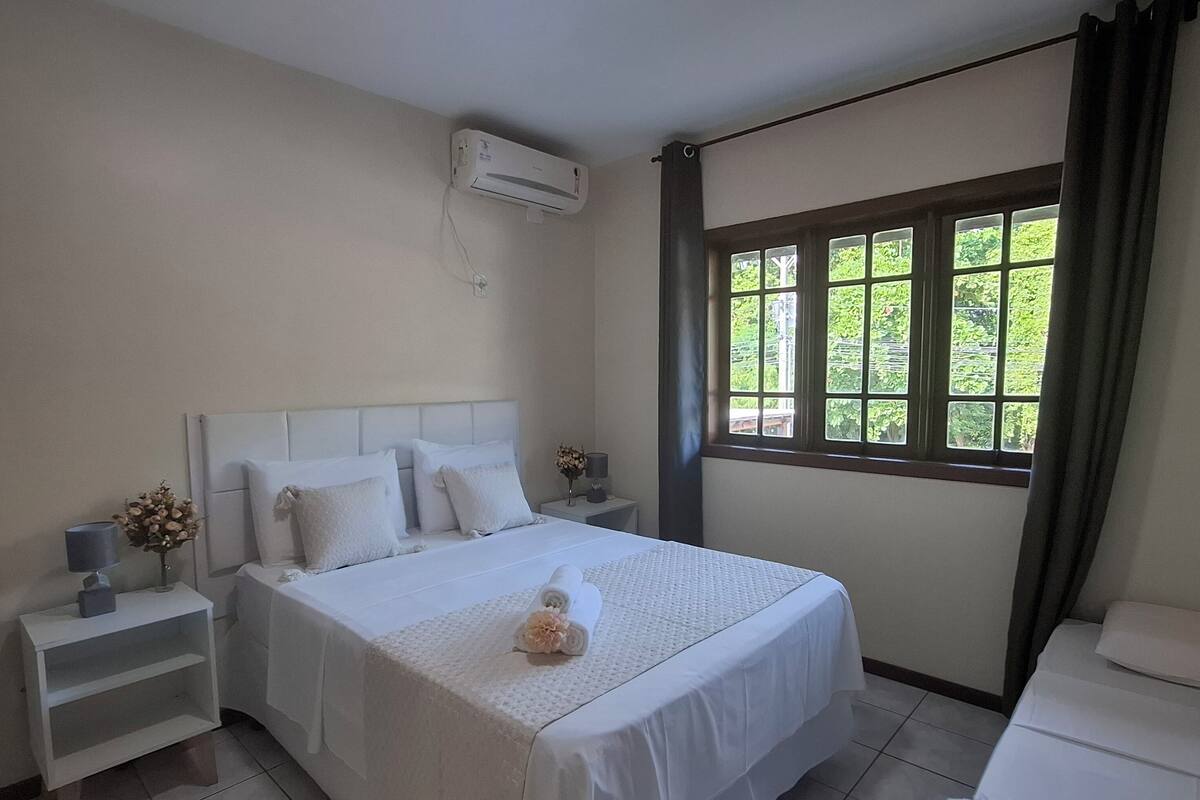 Quarto de hotel com cama, mesas de cabeceira, janela com cortinas e aparelho de ar condicionado.