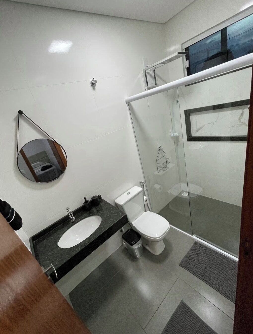 Um banheiro moderno com box de vidro, espelho redondo, vaso sanitário e pia.