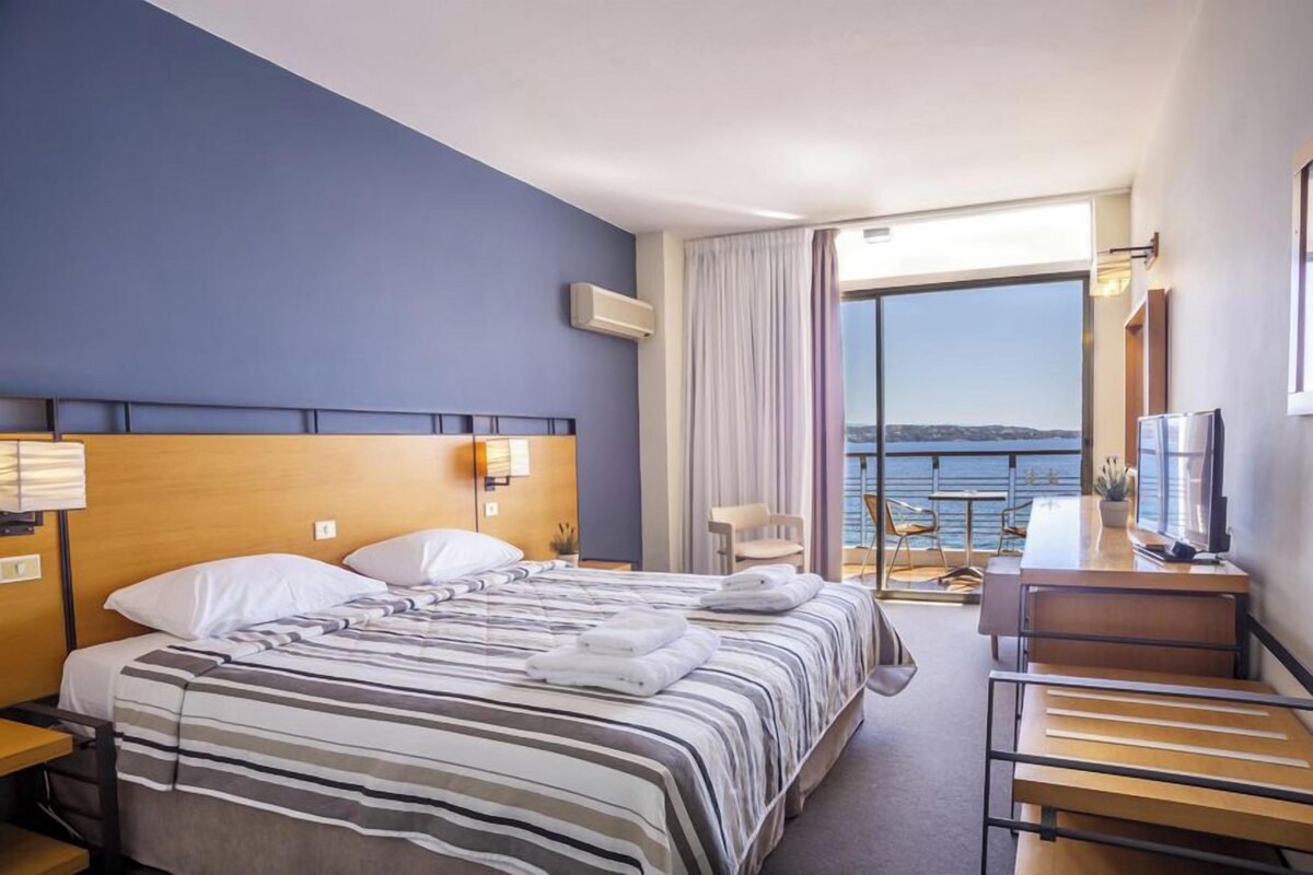 Quarto de hotel com cama, escrivaninha, cadeira, televisão e vista para o mar.