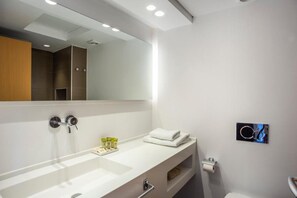 Uma casa de banho moderna com lavatório branco, duas torneiras, um espelho, um porta-toalhas e uma sanita.