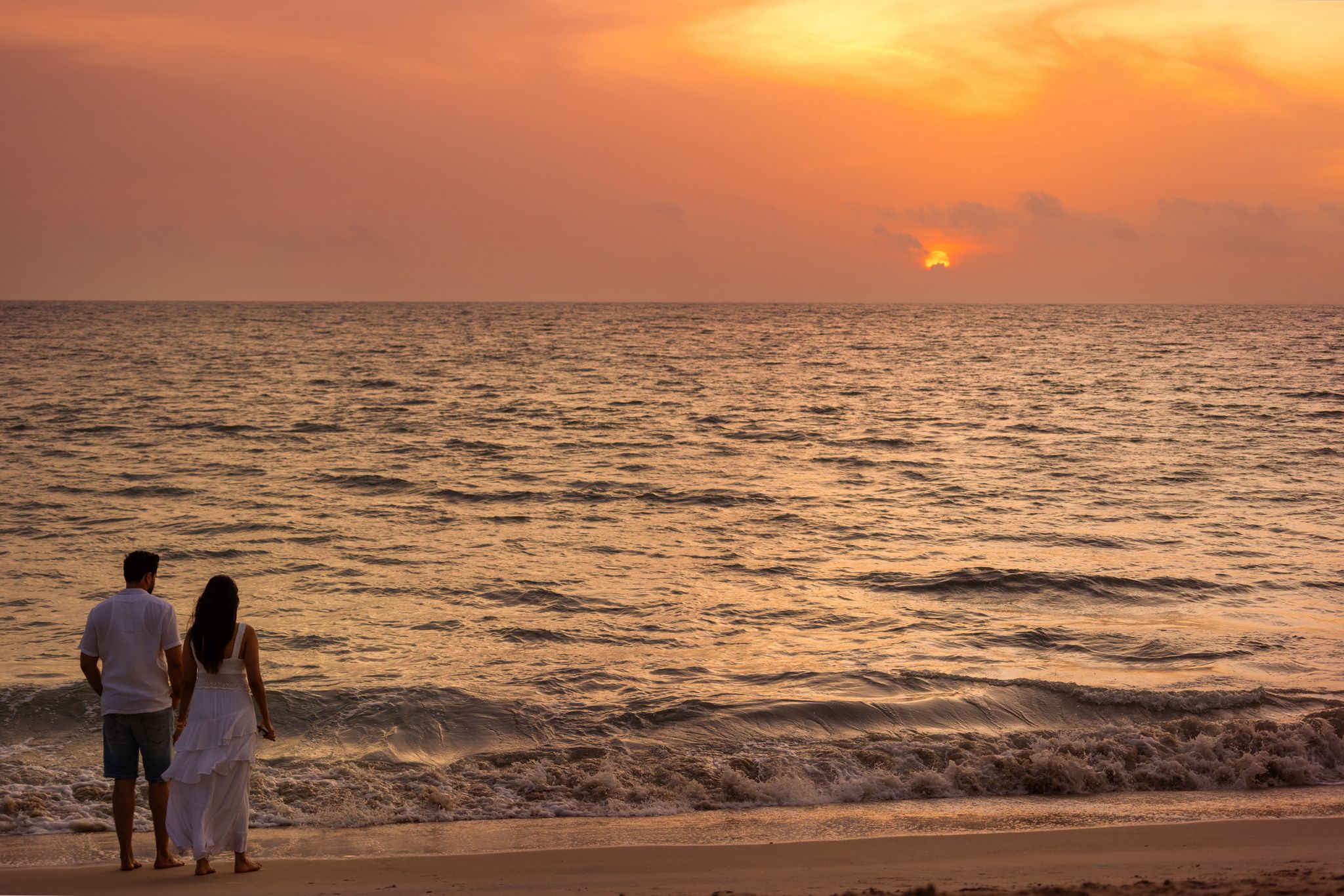 Um casal em pé na praia, de frente para o oceano ao pôr do sol.
