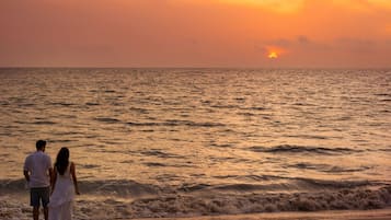 Um casal em pé na praia, de frente para o oceano ao pôr do sol.