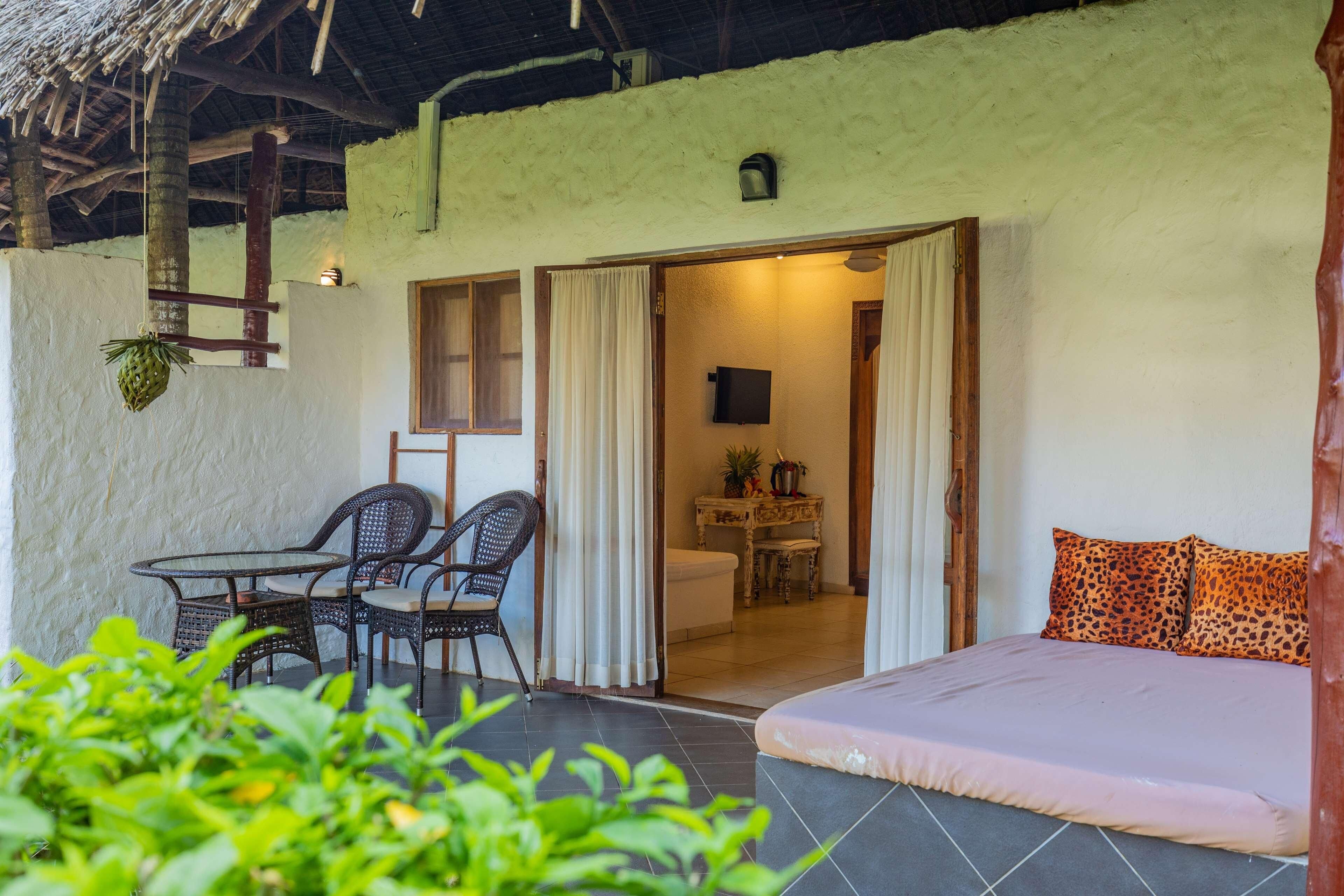 Quarto com uma cama, uma mesa com cadeiras, uma mesinha com uma planta e uma porta que dá para outro cômodo.