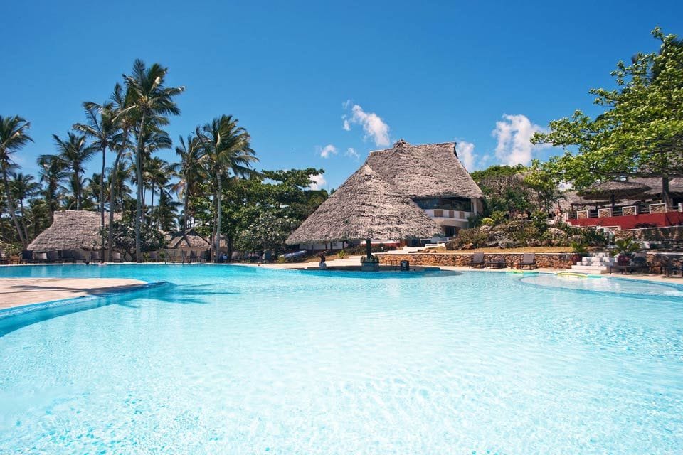 Piscina transparente com um edifício de telhado de palha ao fundo.