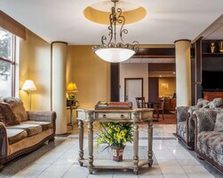 Um saguão de hotel com um lustre clássico, um sofá, uma mesa com um vaso de flores e uma televisão ao fundo.