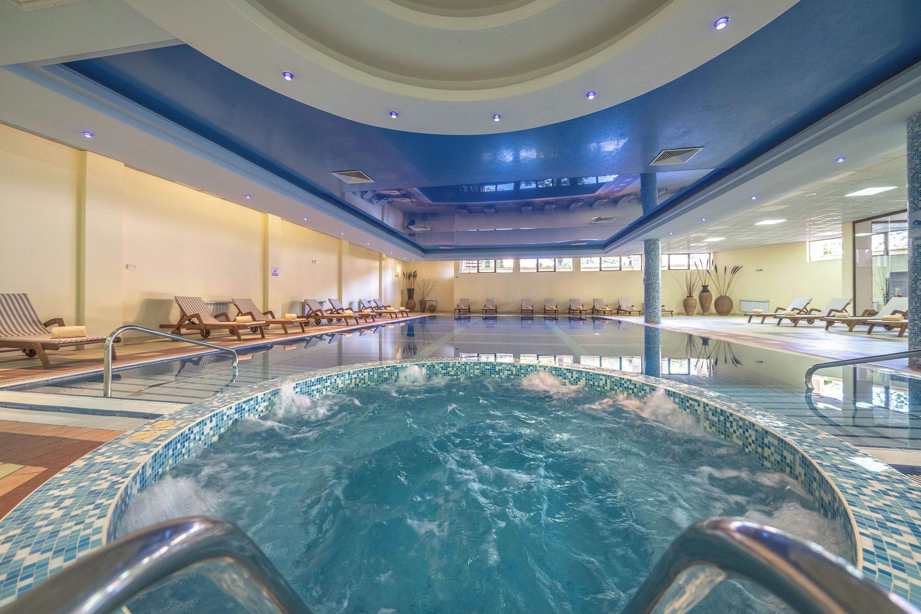 Piscina interior com borda azulejada, espreguiçadeiras e teto com iluminação embutida.
