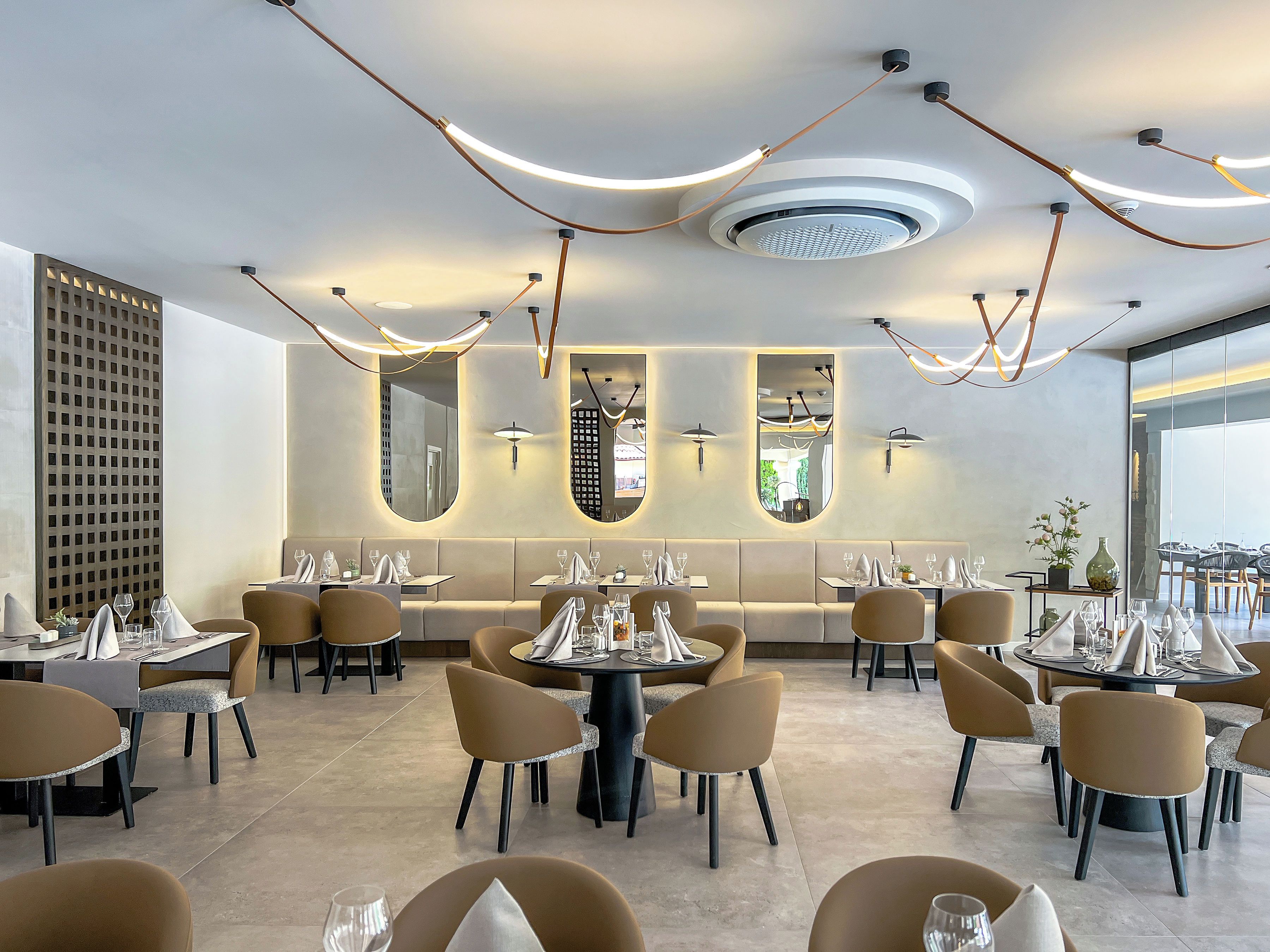 Interior moderno de restaurante com mesas redondas, cadeiras bege e uma área central de estar com um banco acolchoado.