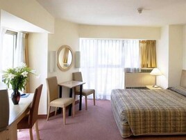 Quarto de hotel com cama, escrivaninha e cadeira, espelho, janela com cortinas e abajur.