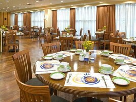 Uma sala de jantar de hotel com mesas redondas preparadas para o pequeno-almoço, cadeiras de madeira e grandes janelas com cortinas.