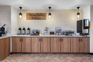 Espaço de café da manhã com uma placa indicando 'Morning Mellow Breakfast', equipado com máquinas de café, uma cesta de frutas e um bebedouro.