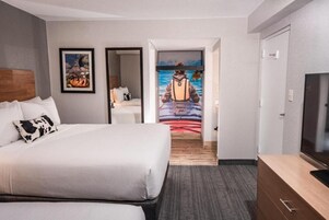 Quarto de hotel com uma cama, televisão, penteadeira e um quadro na parede.