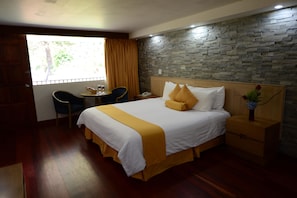 Quarto de hotel com cama, mesa de cabeceira, cadeira, pequena mesa e janela com cortinas.